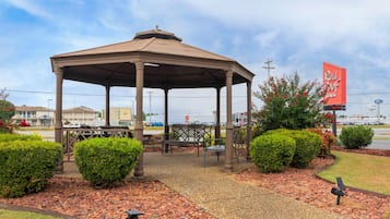 Um gazebo com um banco, rodeado por arbustos bem cuidados e um caminho de pedrinhas.