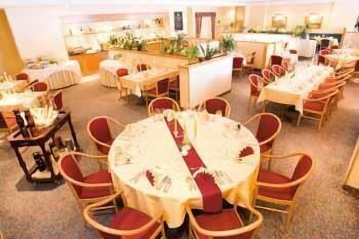 Um espaço de refeições de hotel com mesas redondas e cadeiras vermelhas, uma área de bar e um balcão de recepção.