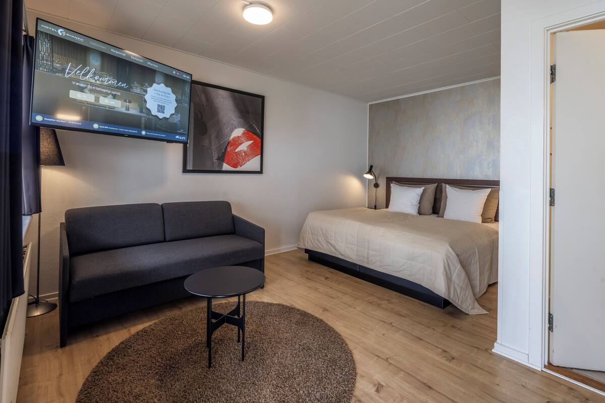 Quarto de hotel moderno com sofá, mesa redonda, cama com cabeceira e televisão fixada na parede exibindo uma mensagem de boas-vindas.