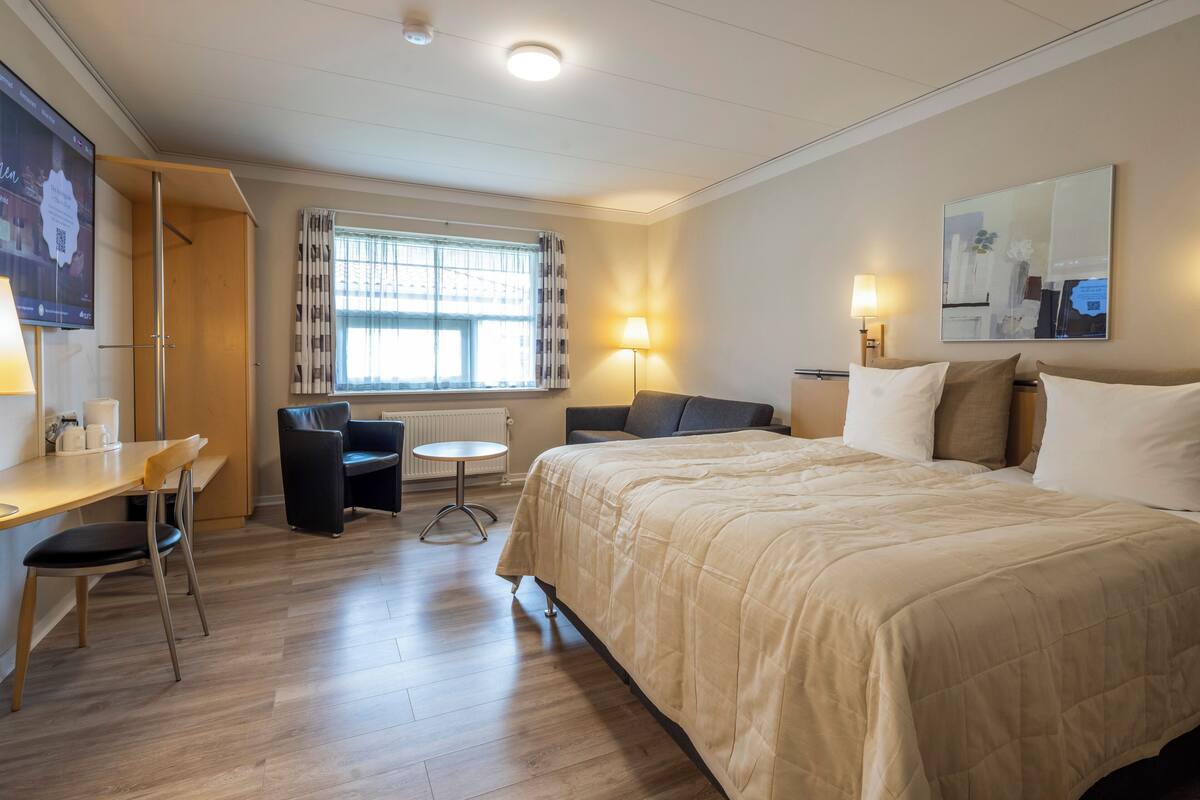 Quarto de hotel com cama, escrivaninha, cadeira, mesa pequena e janela com cortinas.