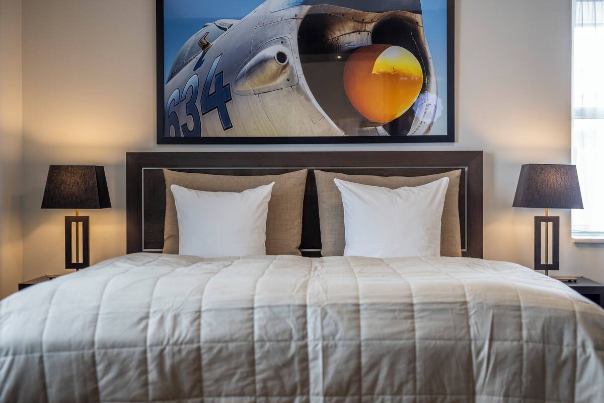 Quarto de hotel com uma cama grande, duas luminárias de cabeceira, uma cabeceira e um quadro emoldurado sobre a cama.