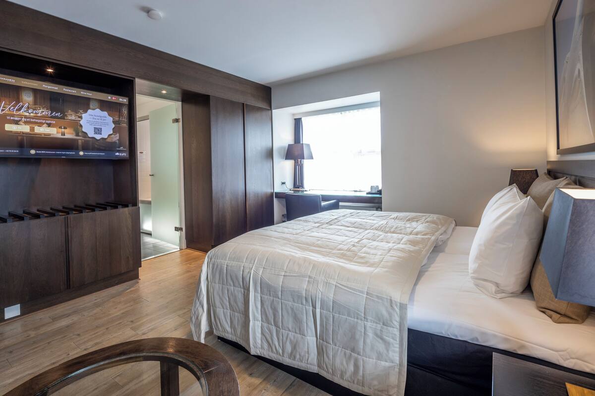 Quarto de hotel moderno com uma cama grande, uma escrivaninha, uma cadeira e uma TV exibindo um menu de restaurante.