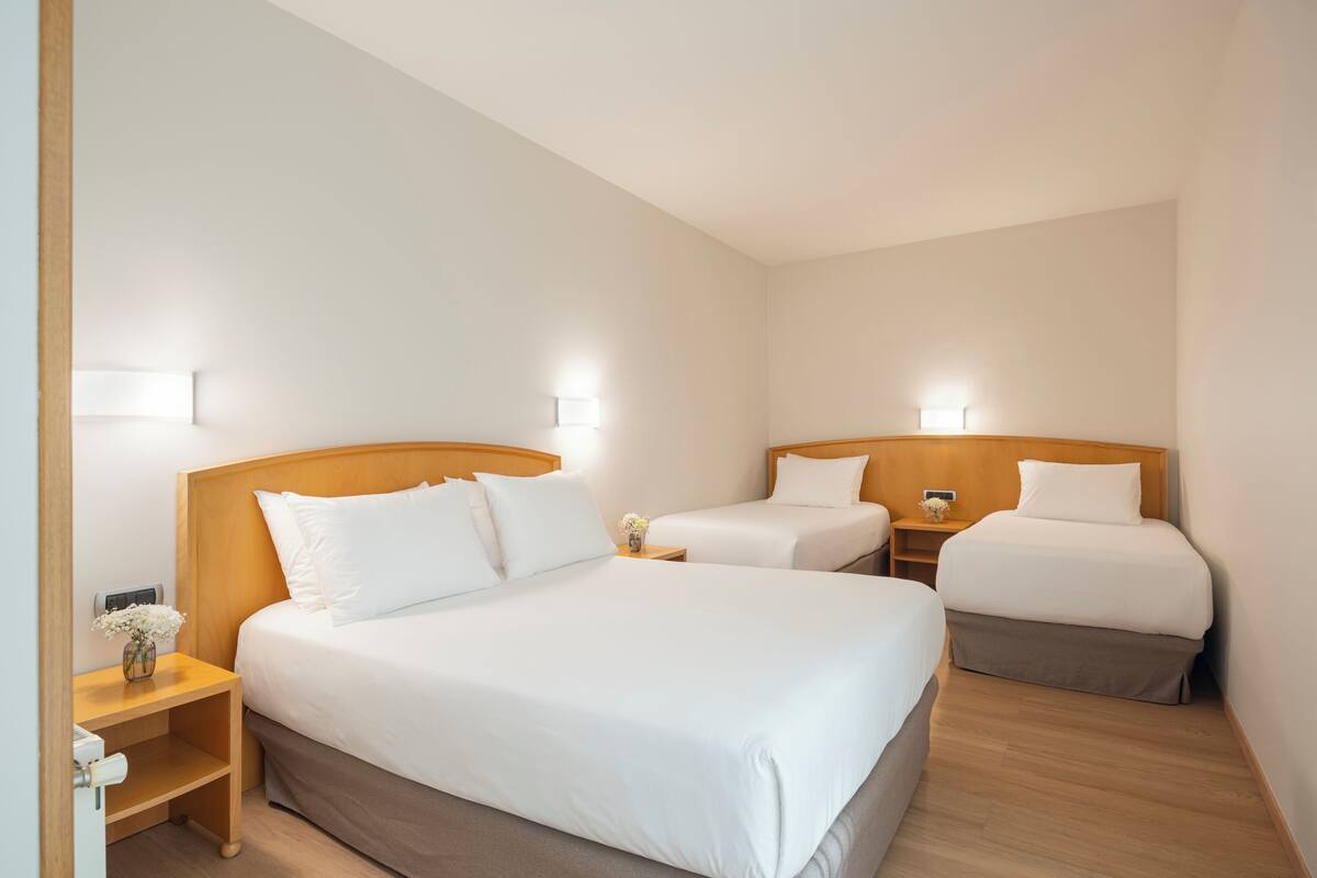 Quarto de hotel com uma cama grande, duas camas menores, cabeceiras de madeira, mesas de cabeceira e luminárias fixadas na parede.