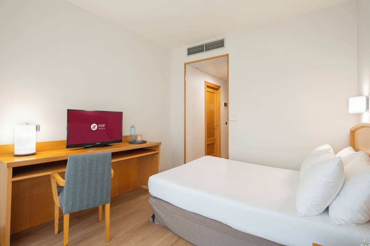 Quarto de hotel com uma cama, uma escrivaninha com um computador, uma cadeira e uma porta.