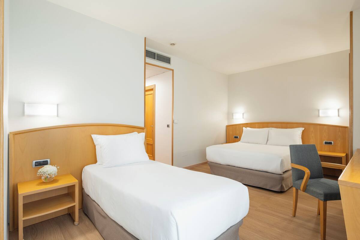 Quarto de hotel com duas camas, uma escrivaninha e uma cadeira. Tem uma mesa de cabeceira com um vaso de flores e um abajur.