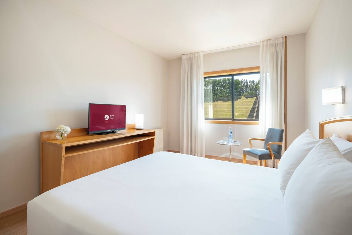 Quarto de hotel com uma cama grande, uma escrivaninha com um laptop, uma cadeira, uma mesinha e uma janela com vista para uma área gramada.
