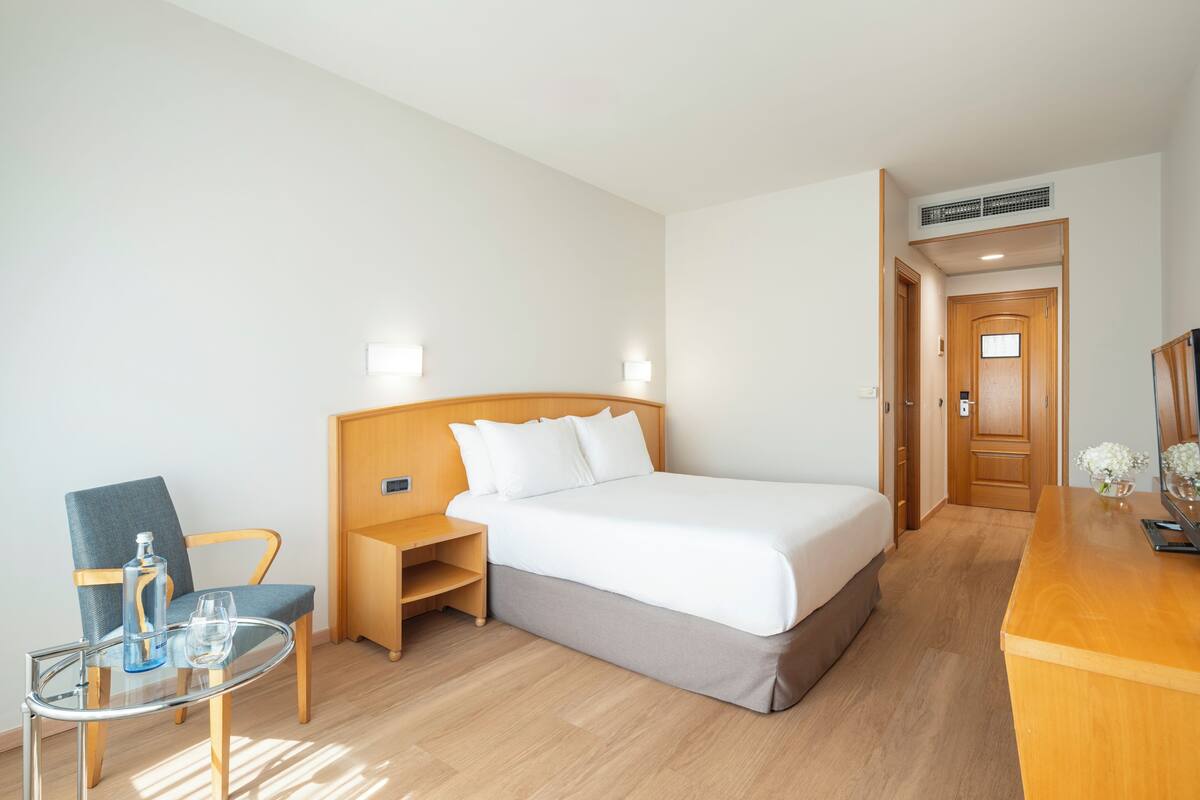 Quarto de hotel com cama, uma cadeira, mesa pequena e escrivaninha de madeira.