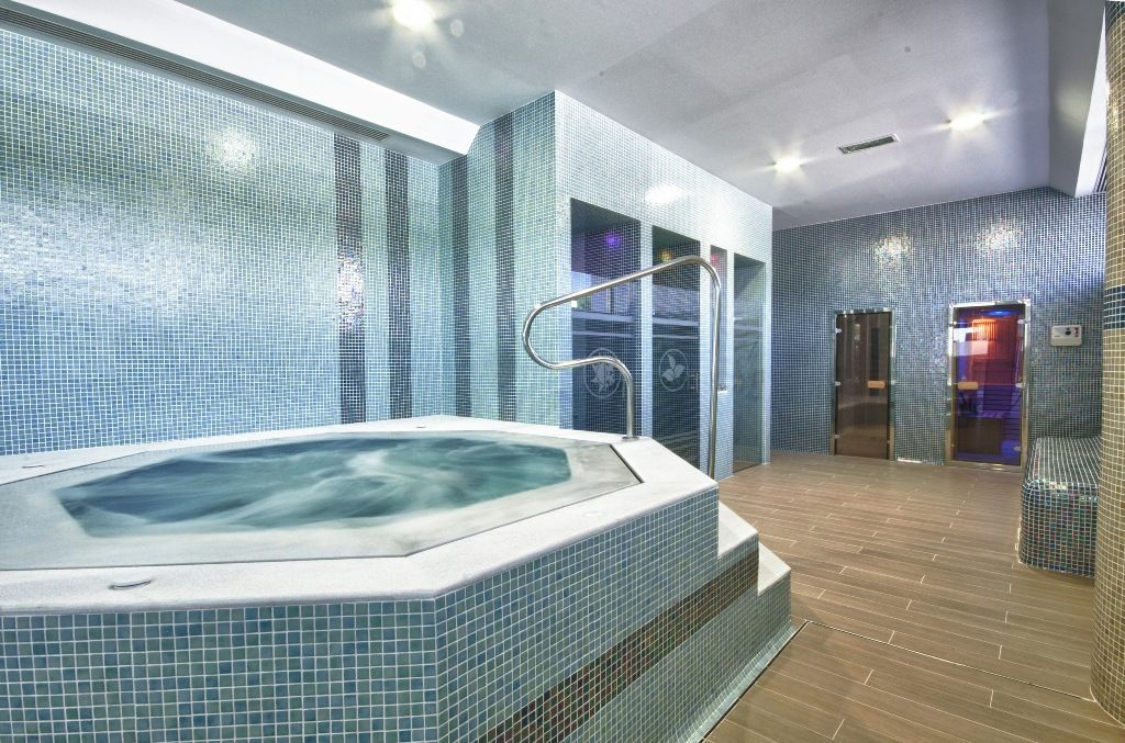 Piscina interior moderna com hidromassagem, paredes azulejadas e piso de madeira.