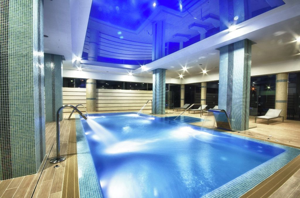 Uma piscina interior com chão em mosaico, área de descanso à beira da piscina e teto com design de iluminação azul.