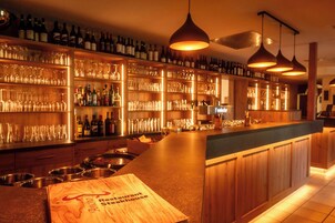 Um bar restaurante com prateleiras de madeira repletas de garrafas e copos, um balcão com cardápio e iluminação pendente.