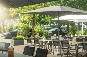Um café ao ar livre com mesas e cadeiras sob guarda-sóis, rodeado por vegetação e carros estacionados.