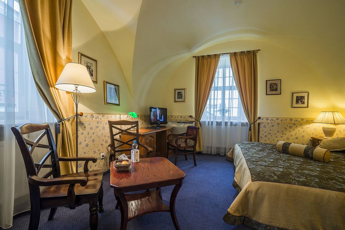 Quarto de hotel com cama, escrivaninha com computador, cadeira e mesa com garrafa e cesta.