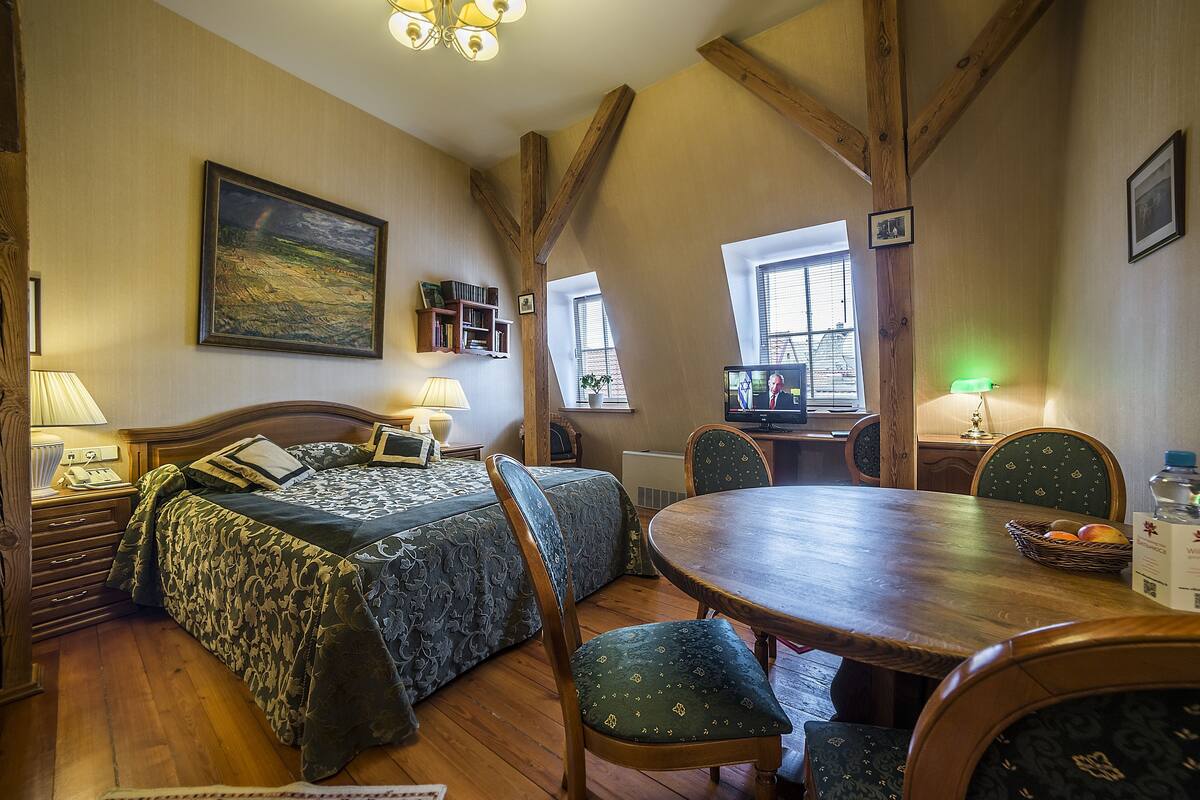 Um quarto aconchegante com uma cama, uma mesa de jantar e cadeiras. Há uma janela com persianas, um abajur e um quadro na parede.