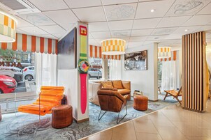 Um saguão de hotel com design moderno, apresentando um sofá de couro, decoração listrada em laranja e branco, e uma grande janela com vista para carros estacionados.