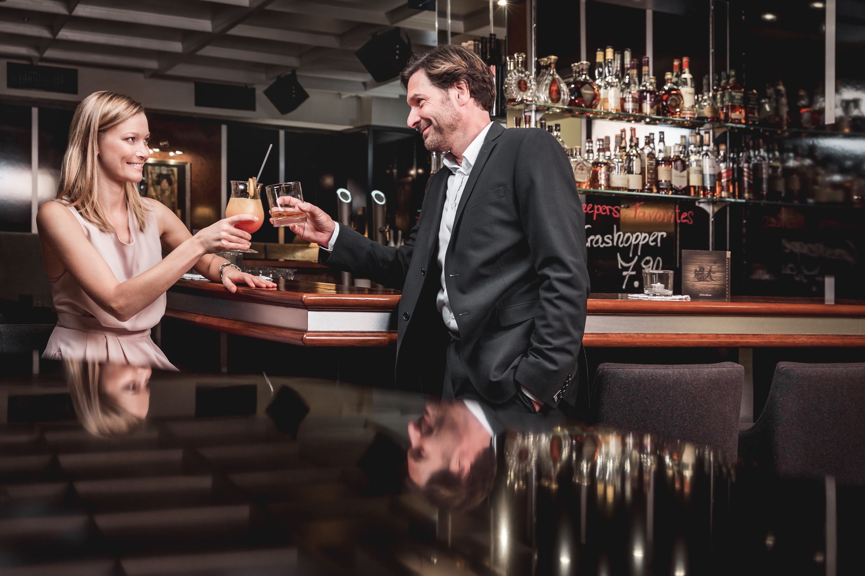 Uma área de bar com um bartender e um cliente. Há prateleiras com garrafas e copos ao fundo.