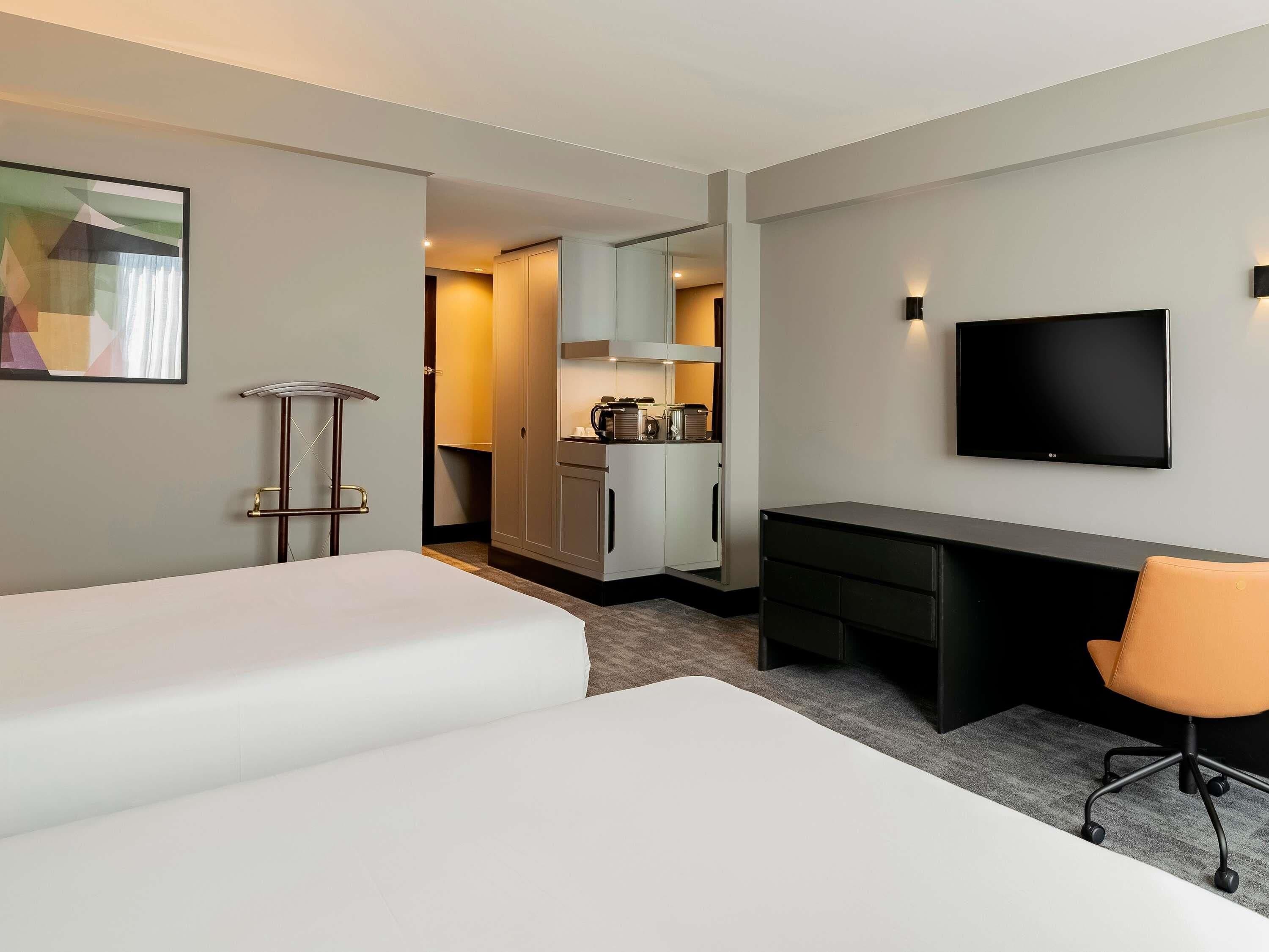Quarto de hotel com cama, escrivaninha, cadeira, televisão e cozinha compacta.