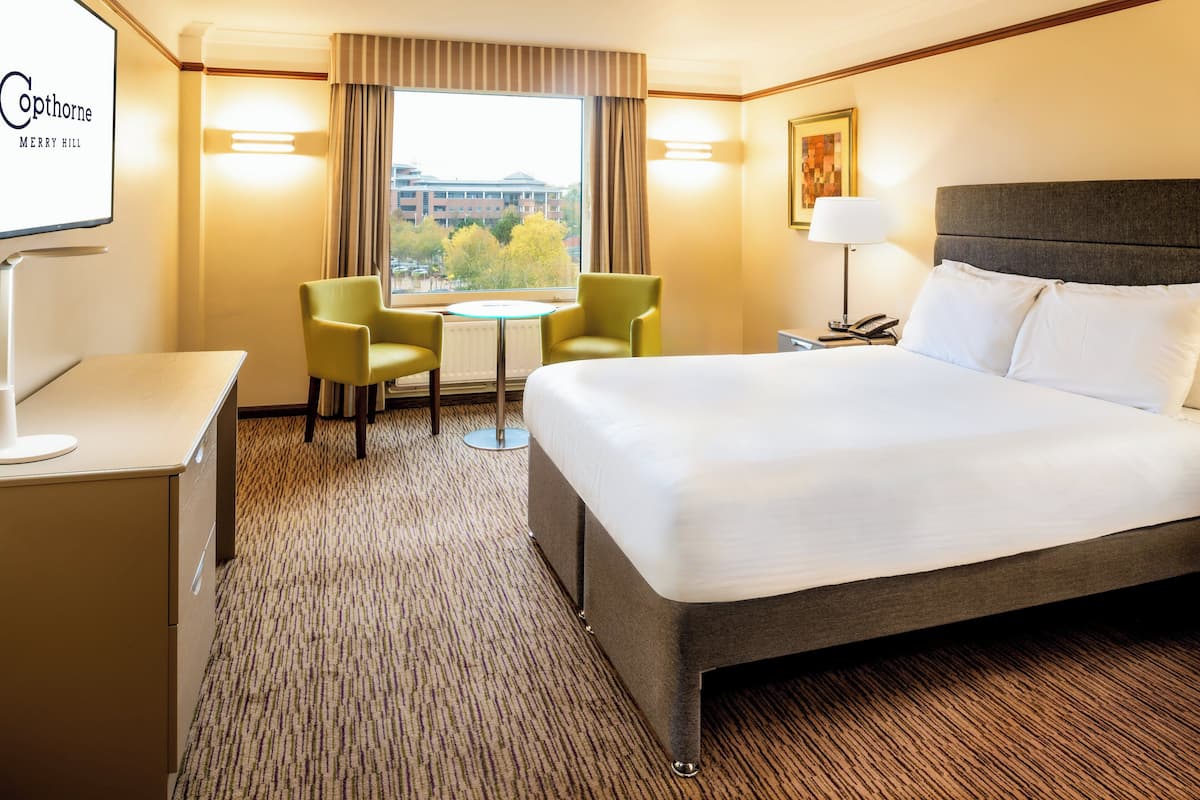 Quarto de hotel com uma cama grande, duas cadeiras, uma mesa pequena e uma televisão exibindo o nome do hotel.