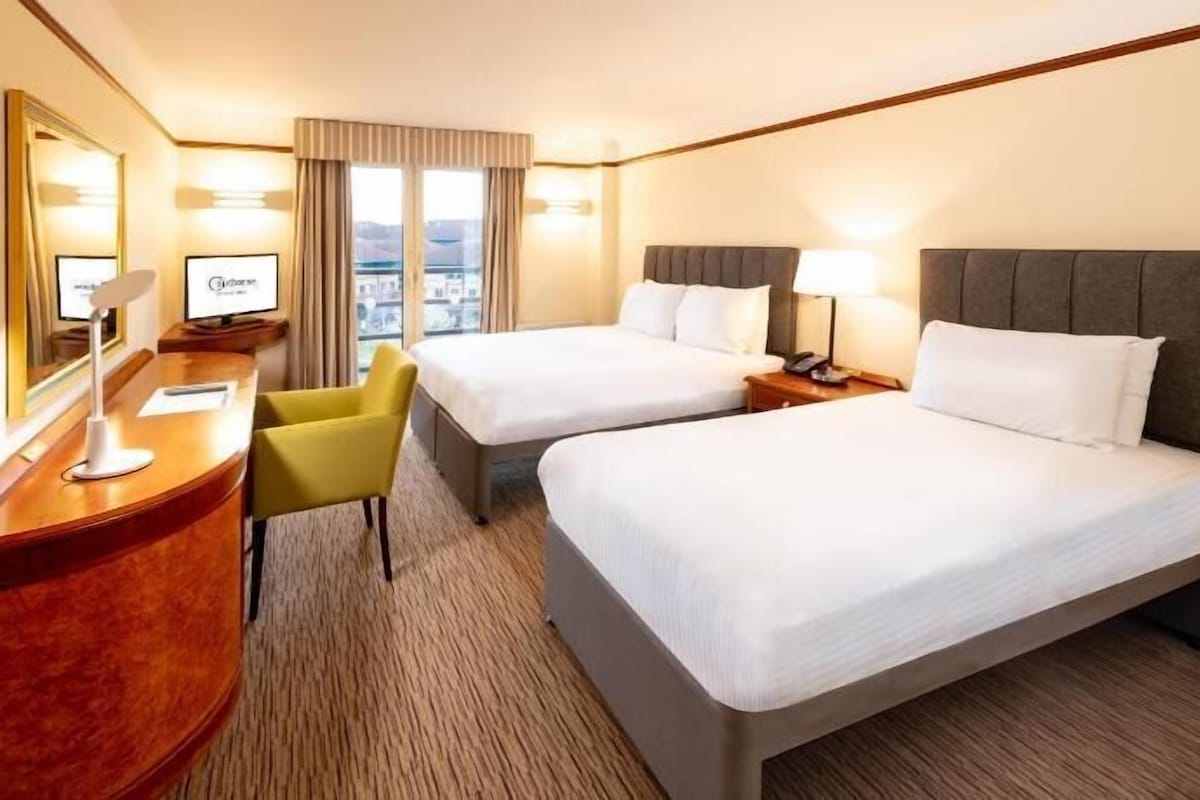 Quarto de hotel com duas camas, uma escrivaninha, uma cadeira, um espelho e uma janela com cortinas.