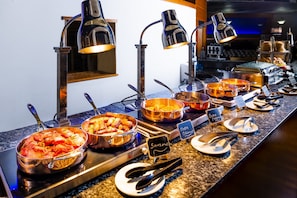 Buffet com diversos pratos e utensílios de servir, incluindo um aviso com a palavra 'Linguiça'.