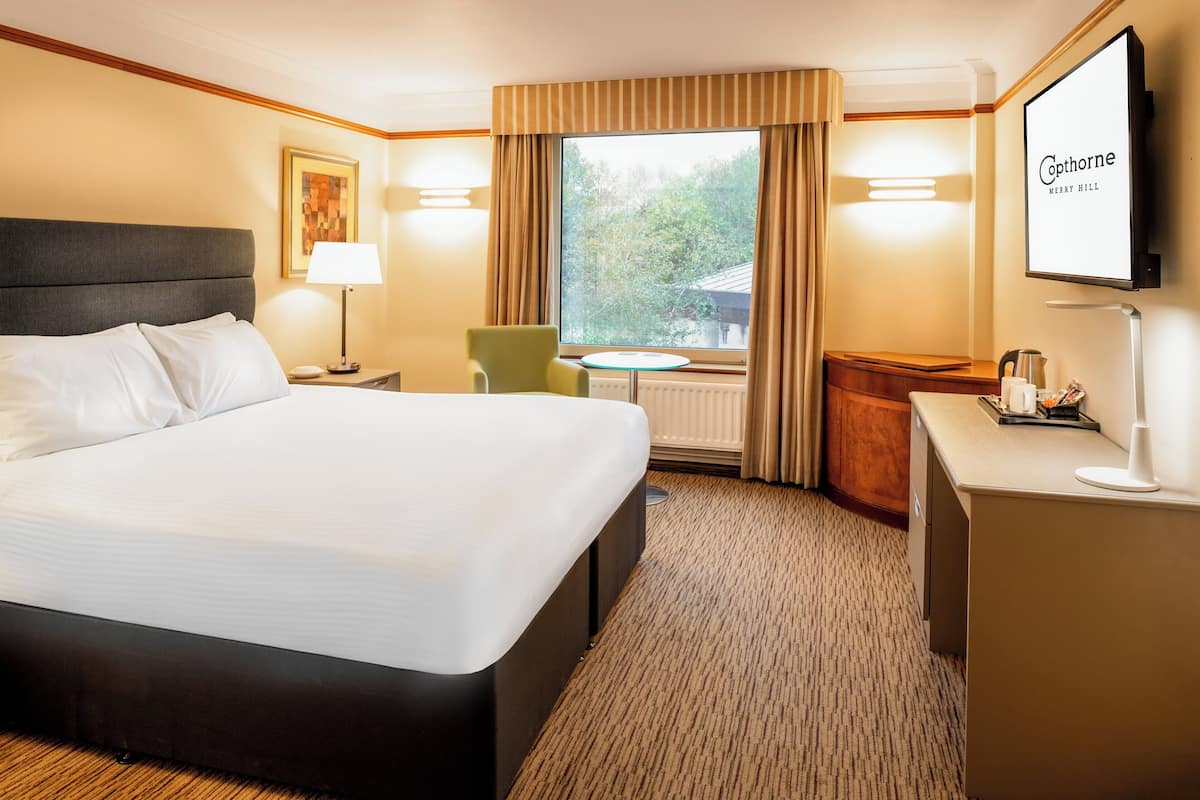 Quarto de hotel com uma cama grande, uma escrivaninha, uma cadeira, uma mesinha e uma televisão na parede.