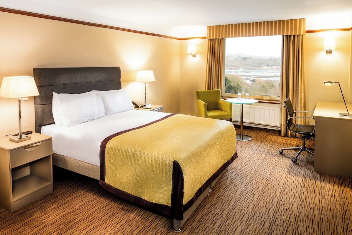 Quarto de hotel com cama, duas mesas de cabeceira, uma cadeira, escrivaninha e janela com vista.