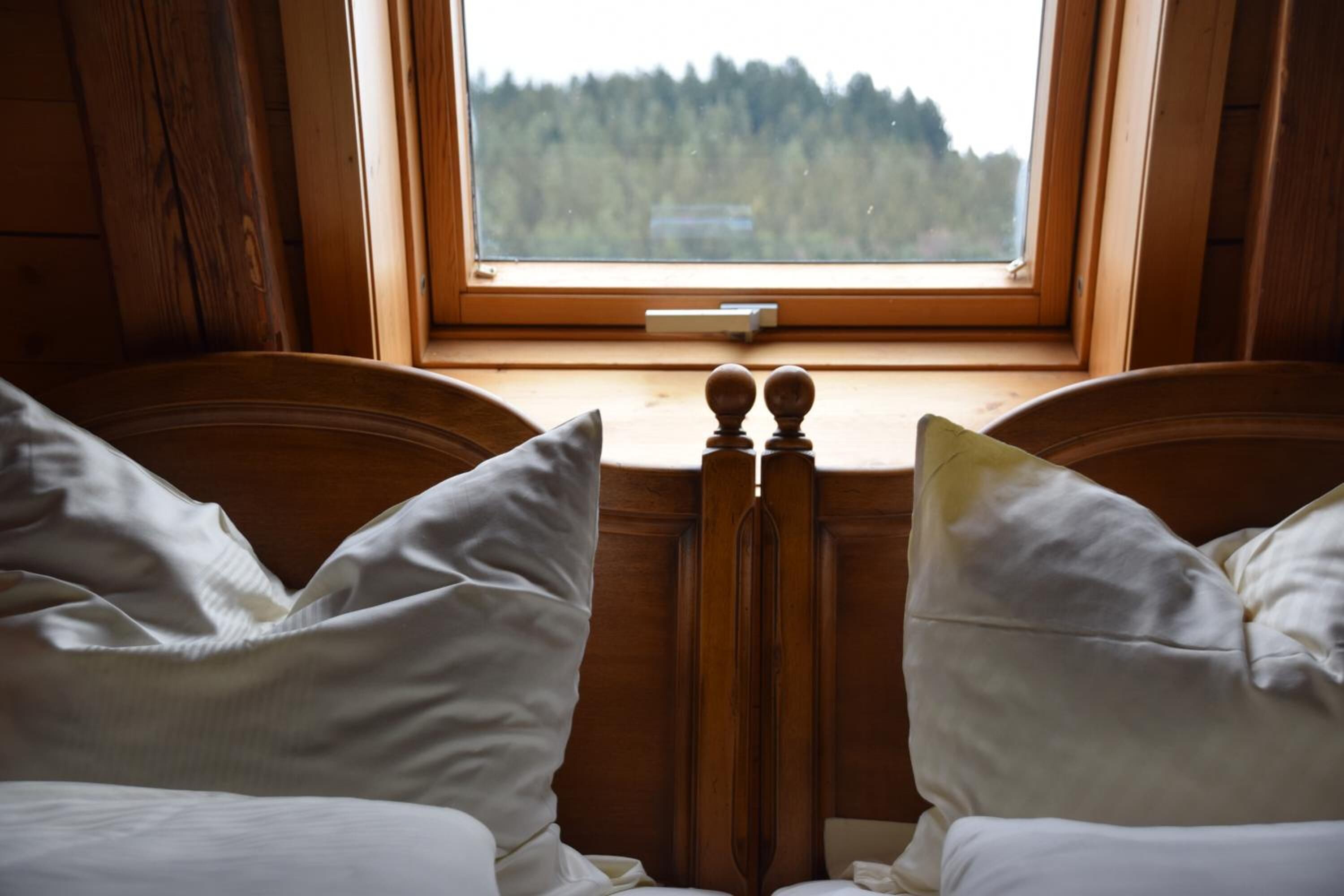 Quarto com duas camas de madeira, uma janela com vista para uma floresta e uma mesinha de cabeceira de madeira entre as camas.