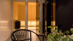Corredor de hotel com uma cadeira e uma planta, que leva a um quarto com cama e janela.