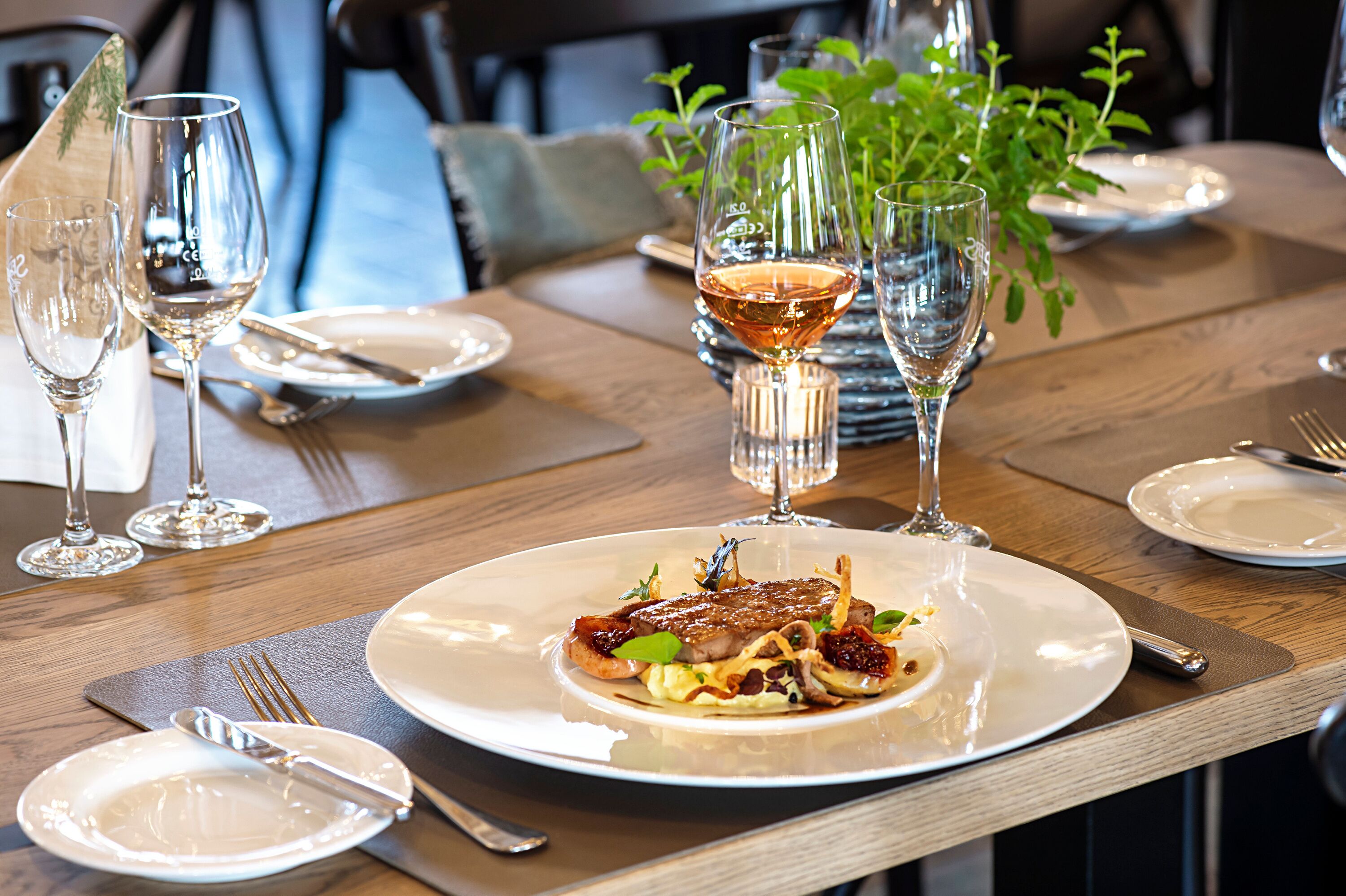 Uma mesa de jantar posta com pratos, talheres e copos de vinho, com uma planta ao fundo.
