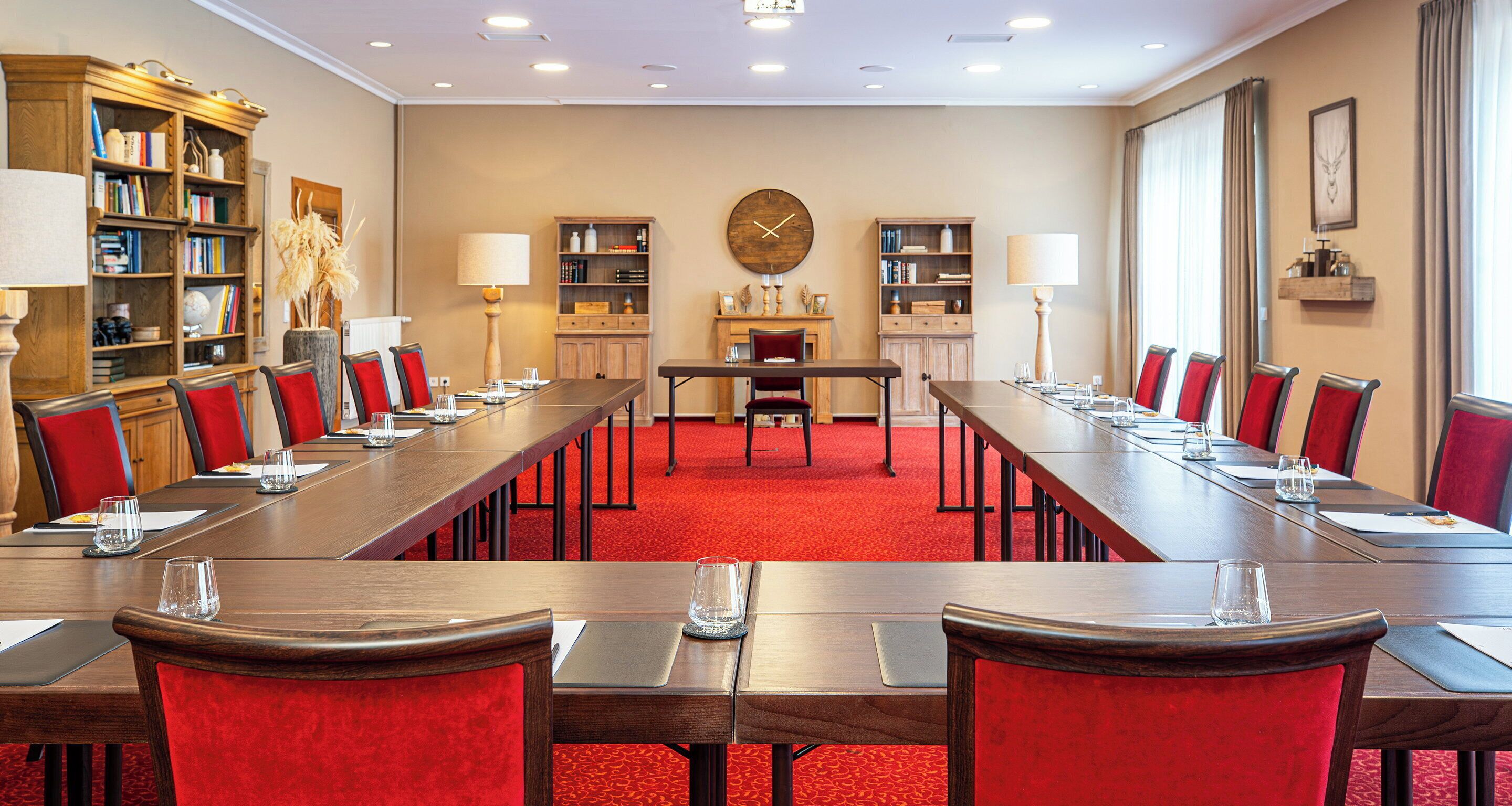 Uma sala de conferências com uma mesa comprida, cadeiras vermelhas, estantes e um relógio na parede.