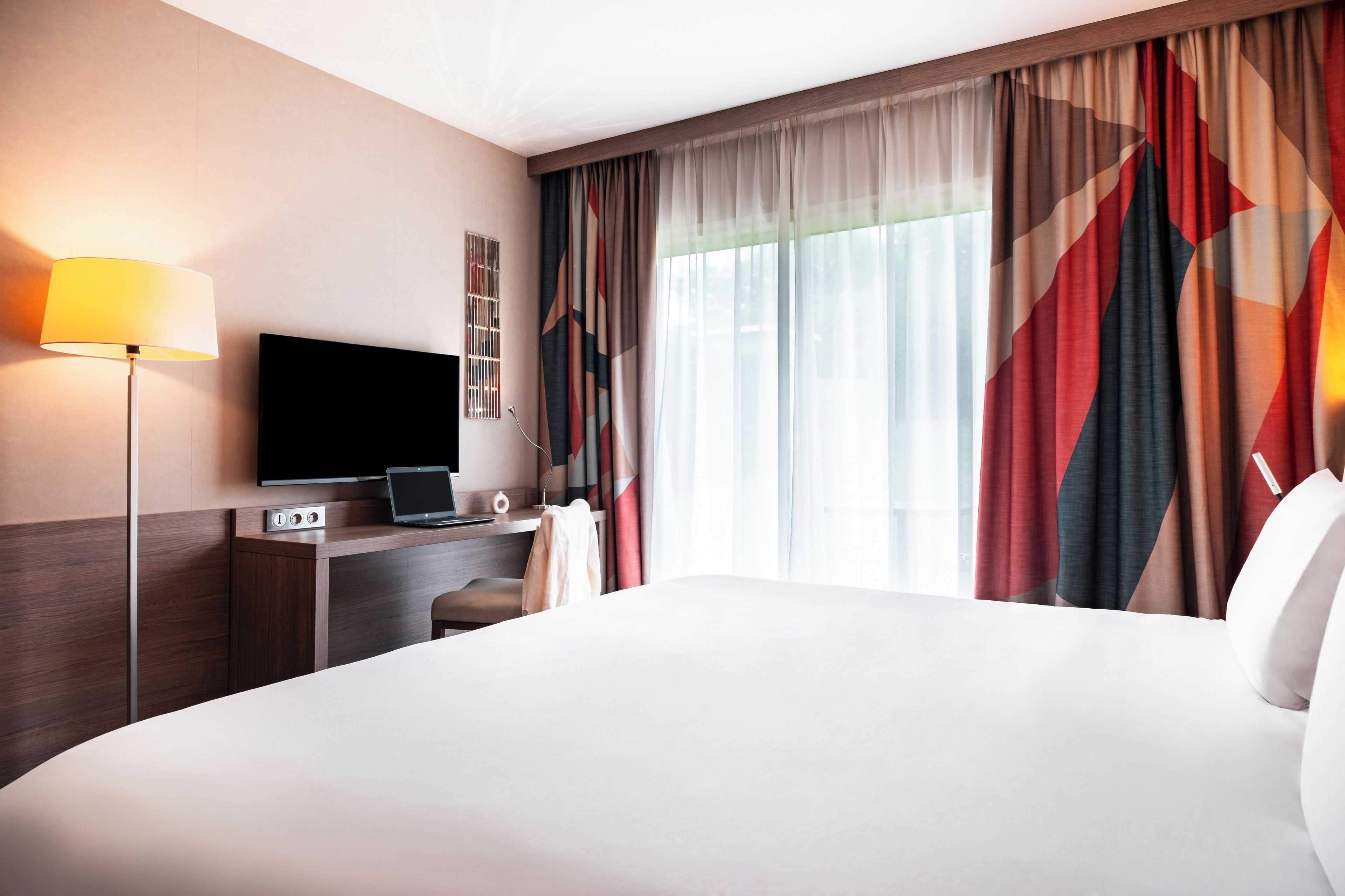 Quarto de hotel com uma cama grande, uma escrivaninha com um laptop, uma televisão de tela plana e um abajur.