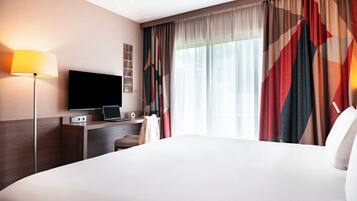 Quarto de hotel com uma cama grande, uma escrivaninha com um laptop, uma televisão de tela plana e um abajur.