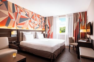 Quarto de hotel com uma cama grande, uma escrivaninha, uma cadeira, uma televisão e uma janela com cortinas.