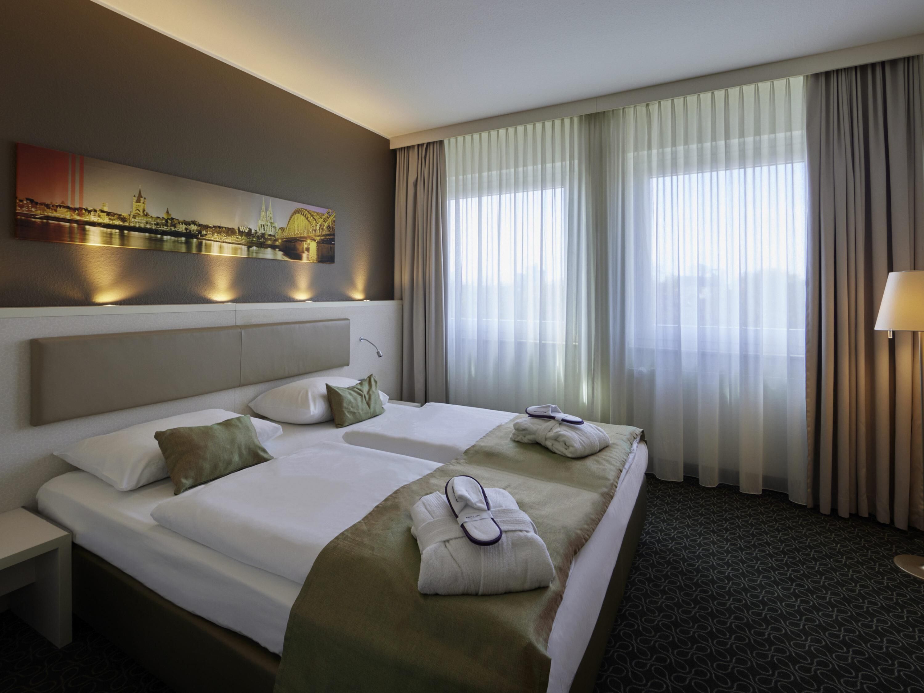 Quarto de hotel com duas camas, cabeceira, mesa de cabeceira, luminária e uma janela grande com cortinas.