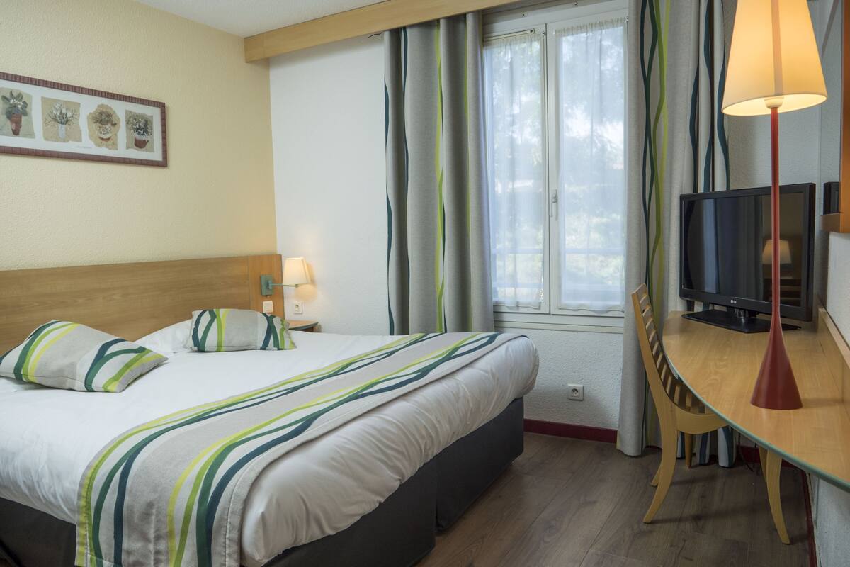 Quarto de hotel com cama, escrivaninha com televisão, luminária e janela com cortinas.