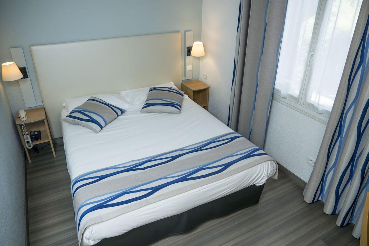 Cama bem arrumada com colcha listrada, duas mesas de cabeceira com abajures, um telefone e uma janela com cortinas.