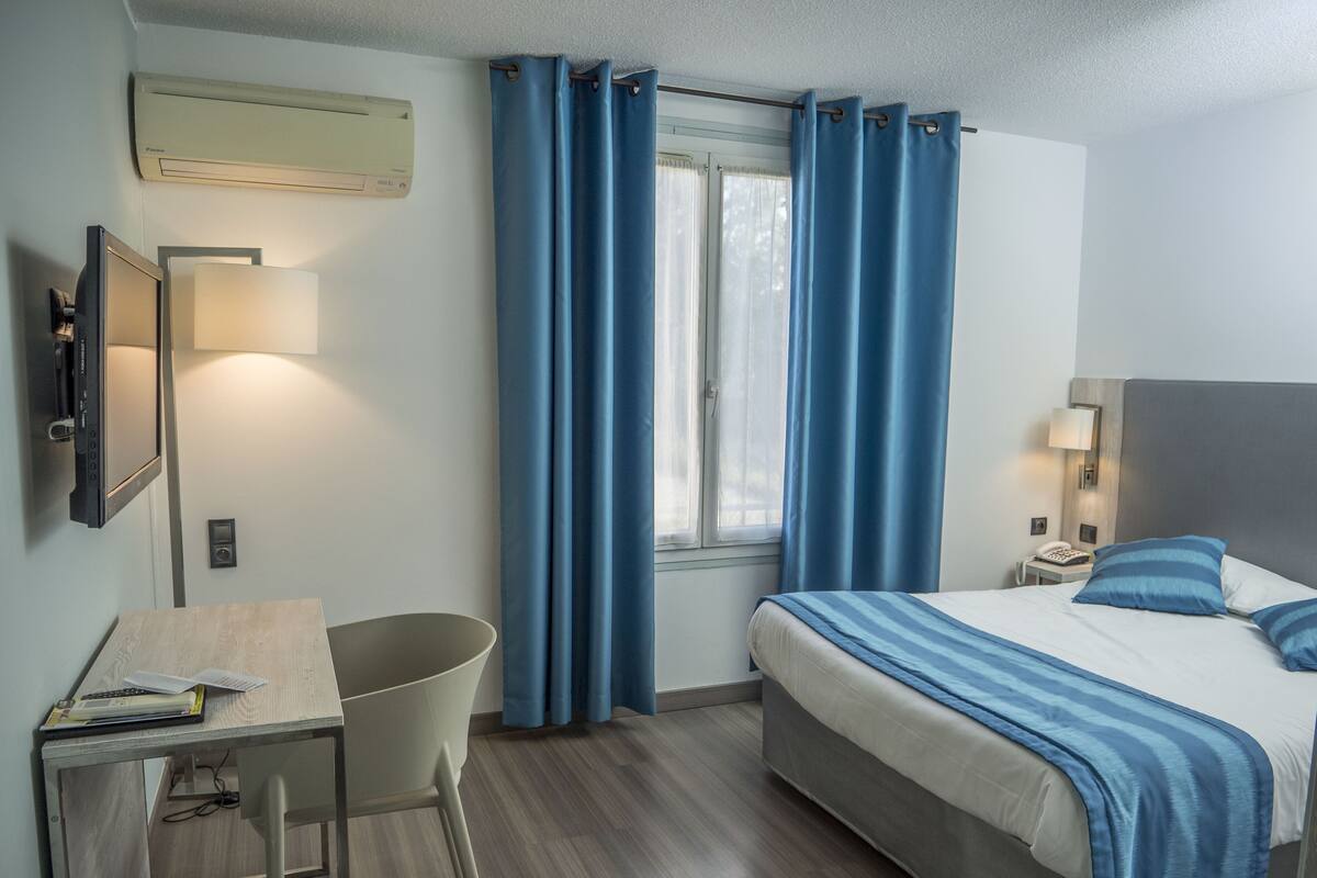 Quarto de hotel com cama, escrivaninha, cadeira e janela com cortinas.