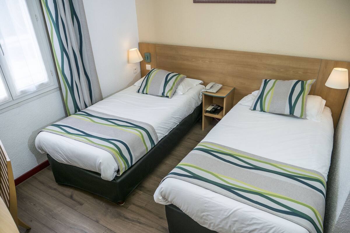 Quarto de hotel com duas camas de solteiro, cada uma com roupa de cama listrada, cabeceira de madeira, luminárias laterais, mesa de cabeceira pequena com telefone e uma janela com cortinas.