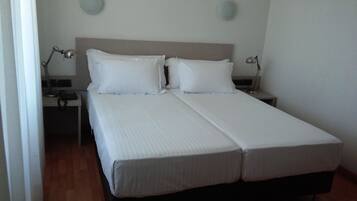 Uma cama de casal com roupa de cama branca, duas mesas de cabeceira com luminárias e piso de madeira.