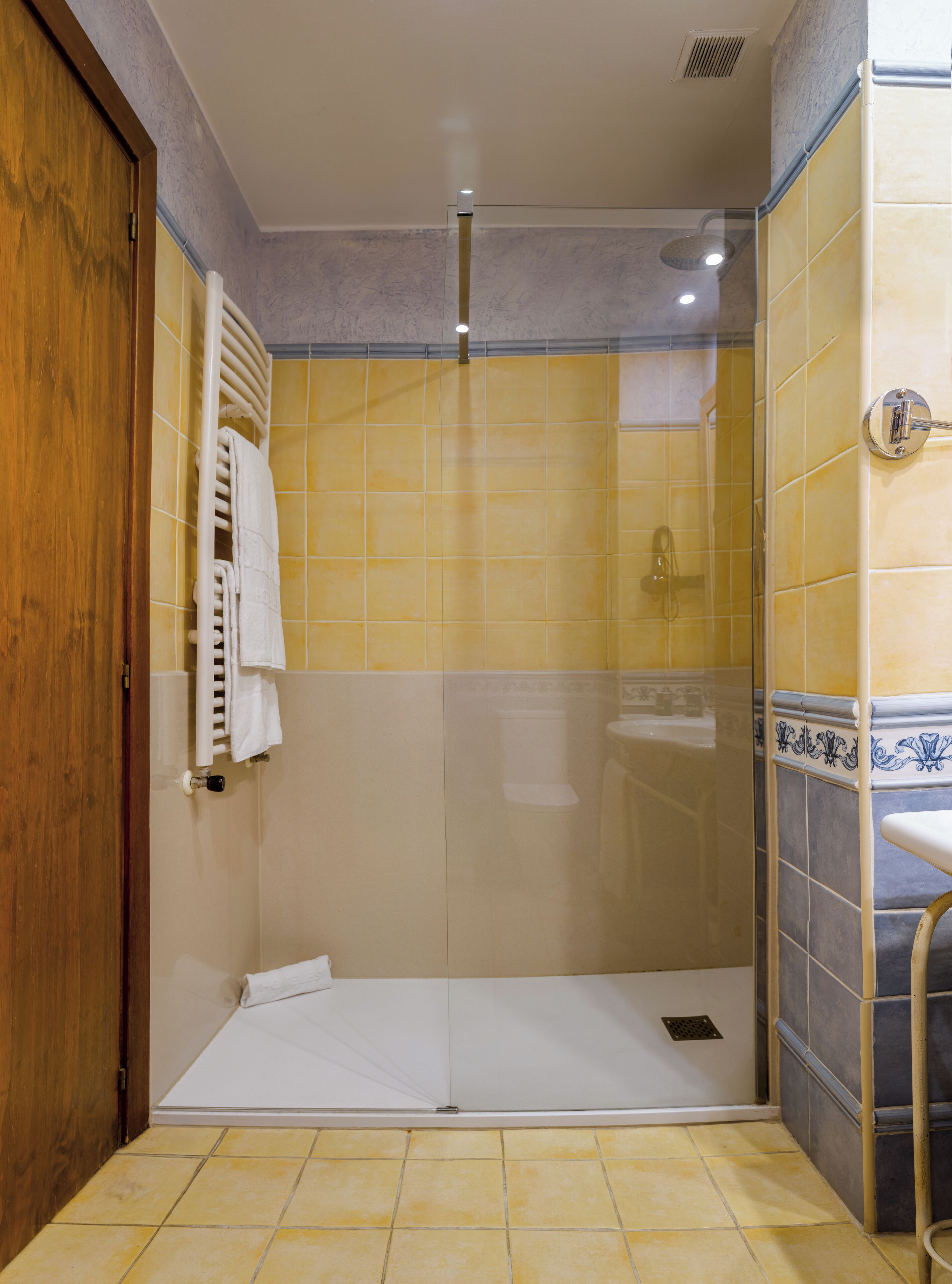 Uma área de chuveiro com azulejos amarelos e beges, uma porta de vidro e uma porta de madeira.