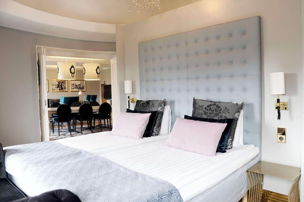 Quarto de hotel moderno com uma cama grande, uma escrivaninha e uma área de jantar visível através de uma porta aberta.