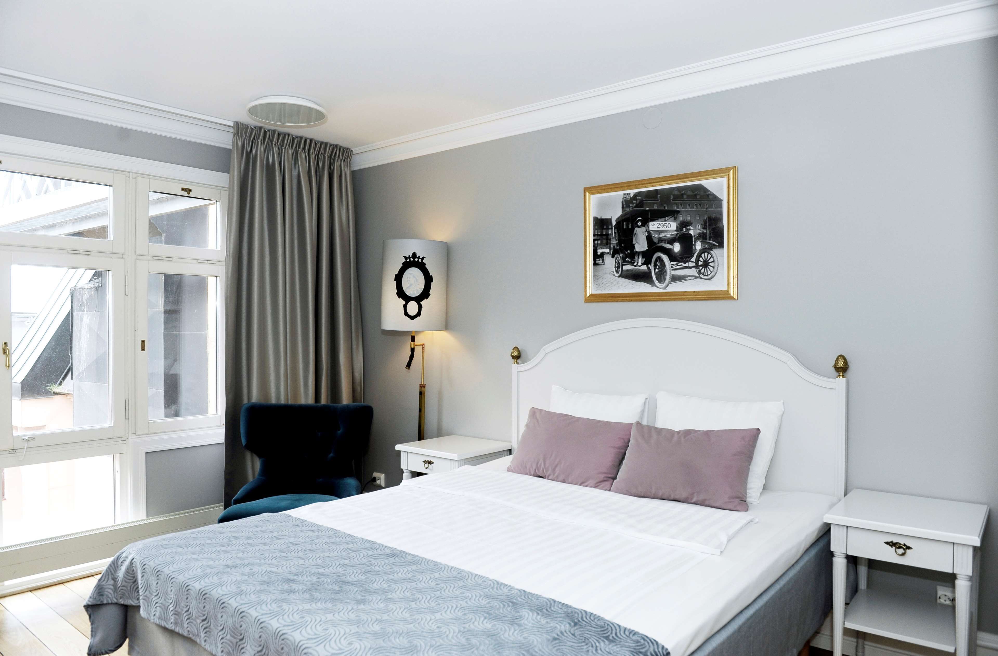 Quarto bem arrumado com uma cama grande, uma cadeira, mesa de cabeceira com abajur, quadro emoldurado na parede e janela com cortinas.