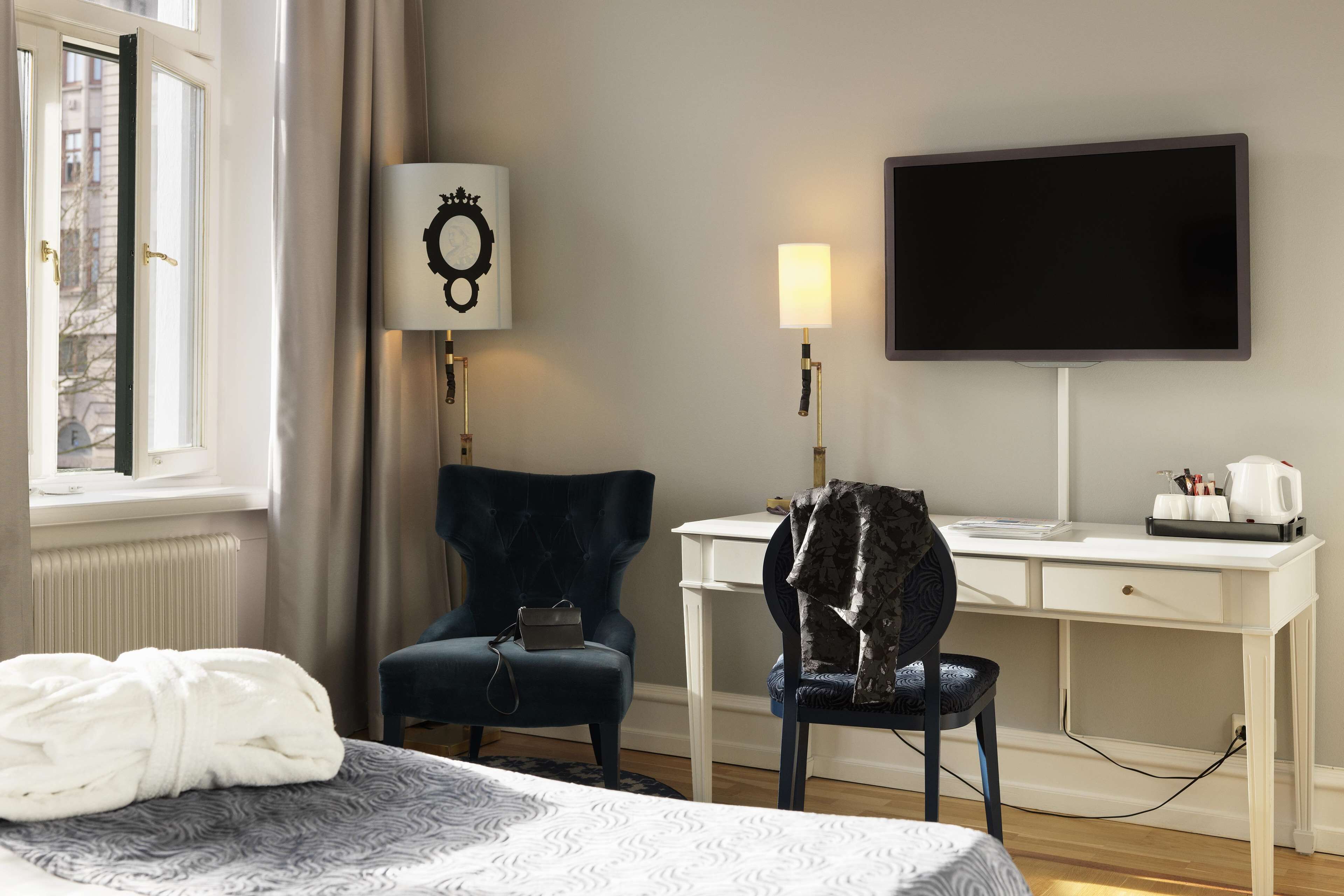 Quarto de hotel com cama, escrivaninha, cadeira, televisão e janela com cortinas.