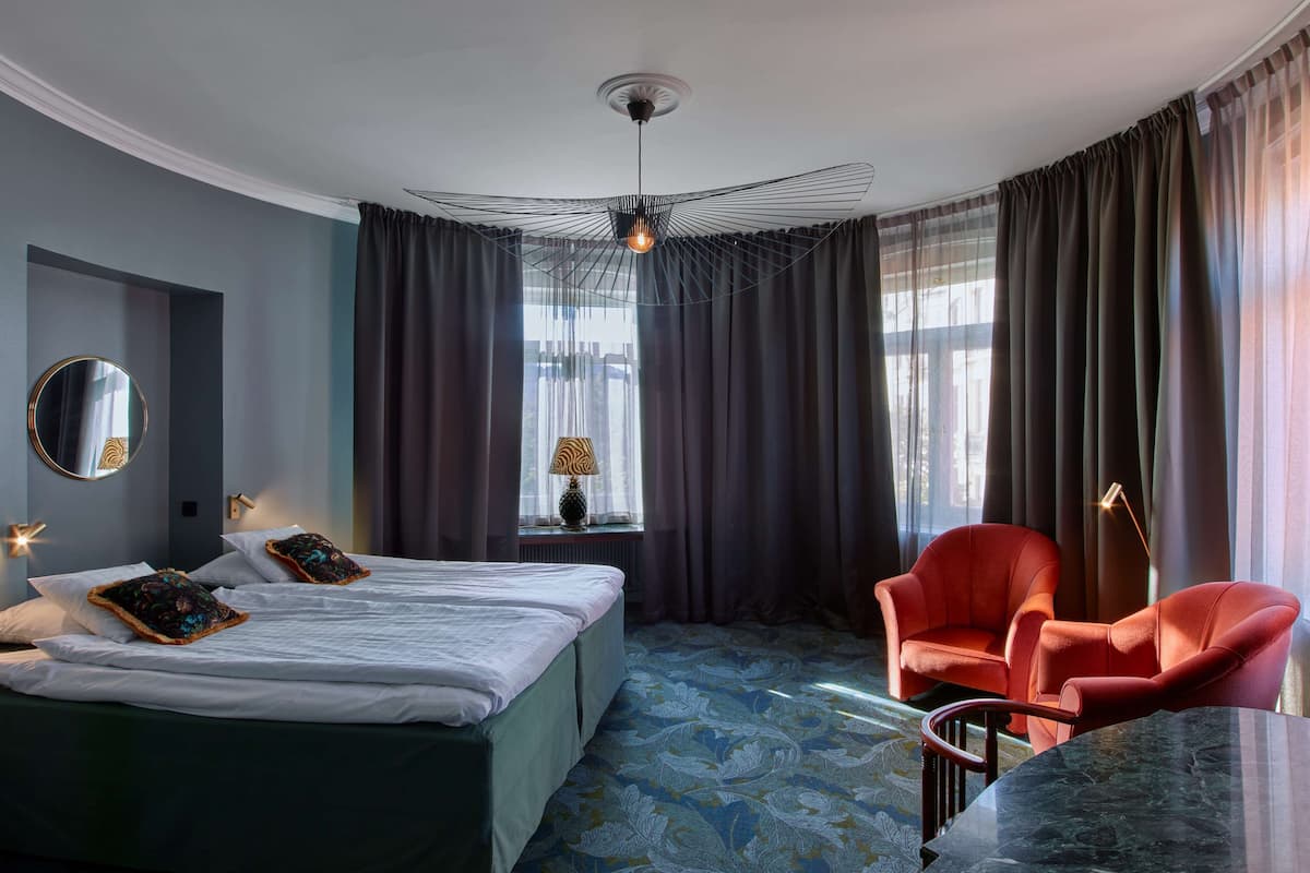 Quarto de hotel com uma cama, duas almofadas, uma poltrona vermelha, um espelho redondo, uma luminária e vista para o exterior.
