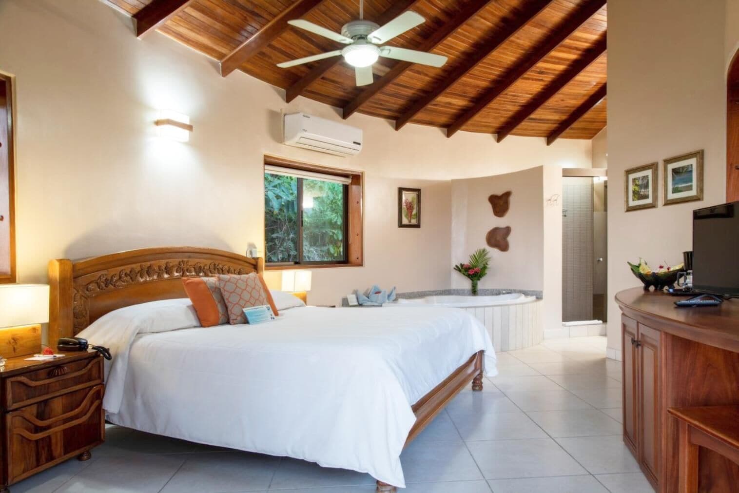 Quarto com teto de madeira, cama com roupa de cama branca, mesa de cabeceira, escrivaninha, cadeira, janela, aparelho de ar condicionado, banheiro com banheira e televisão.