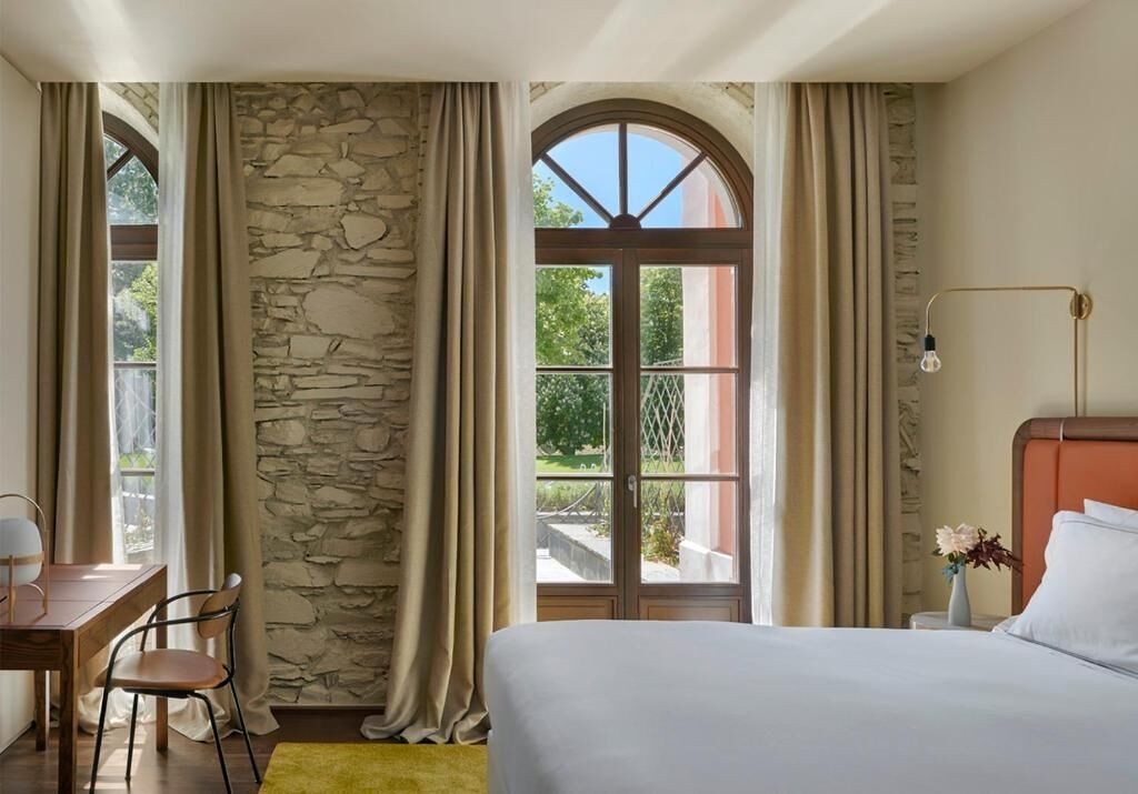 Quarto de hotel com cama, escrivaninha, cadeira, parede de pedra e uma janela grande com cortinas.
