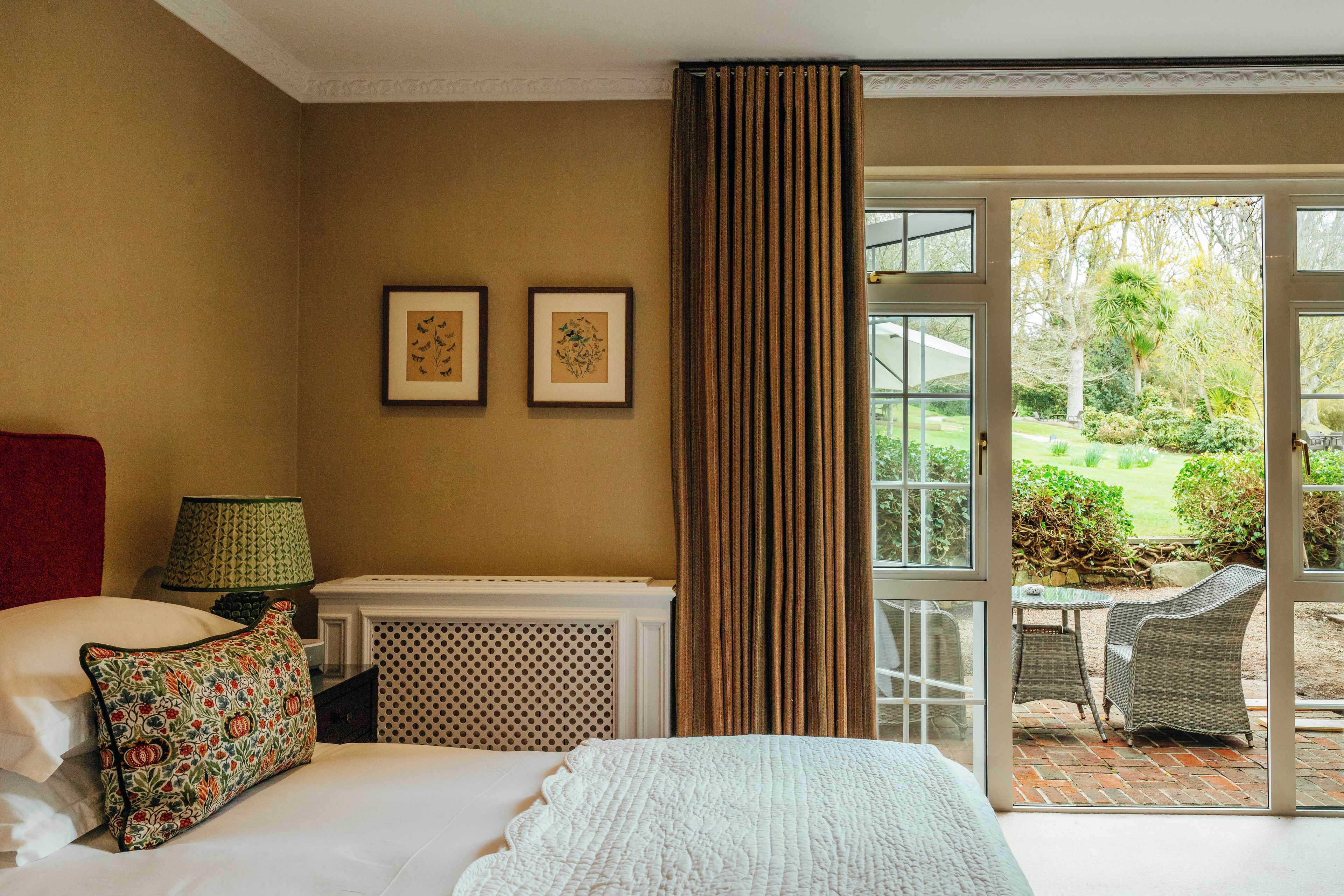 Quarto de hotel com cama, mesa de cabeceira, luminária, duas fotos emolduradas, janela com cortinas e vista para o jardim.
