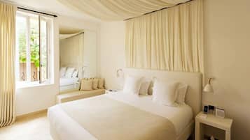 Uma cama grande com lençóis brancos, uma mesinha de cabeceira com abajur e uma janela com cortinas.
