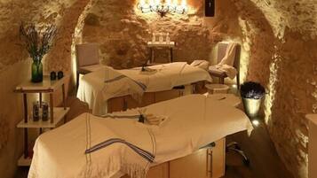 Um quarto acolhedor, com paredes de pedra, duas camas de madeira, uma mesa pequena e um candelabro.