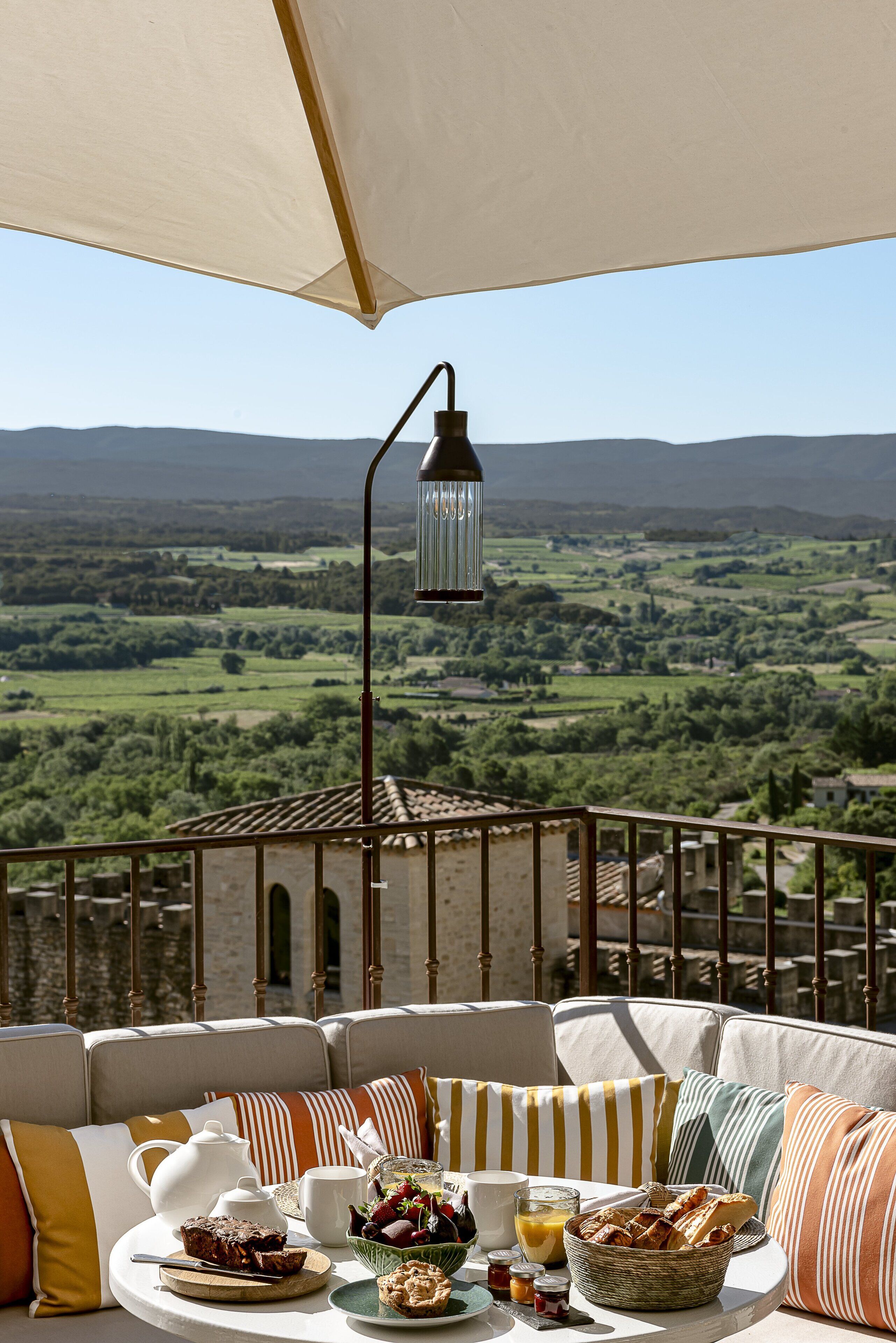 Varanda com mesa posta para o café da manhã, incluindo pão, frutas e bebidas, com vista para uma paisagem deslumbrante.