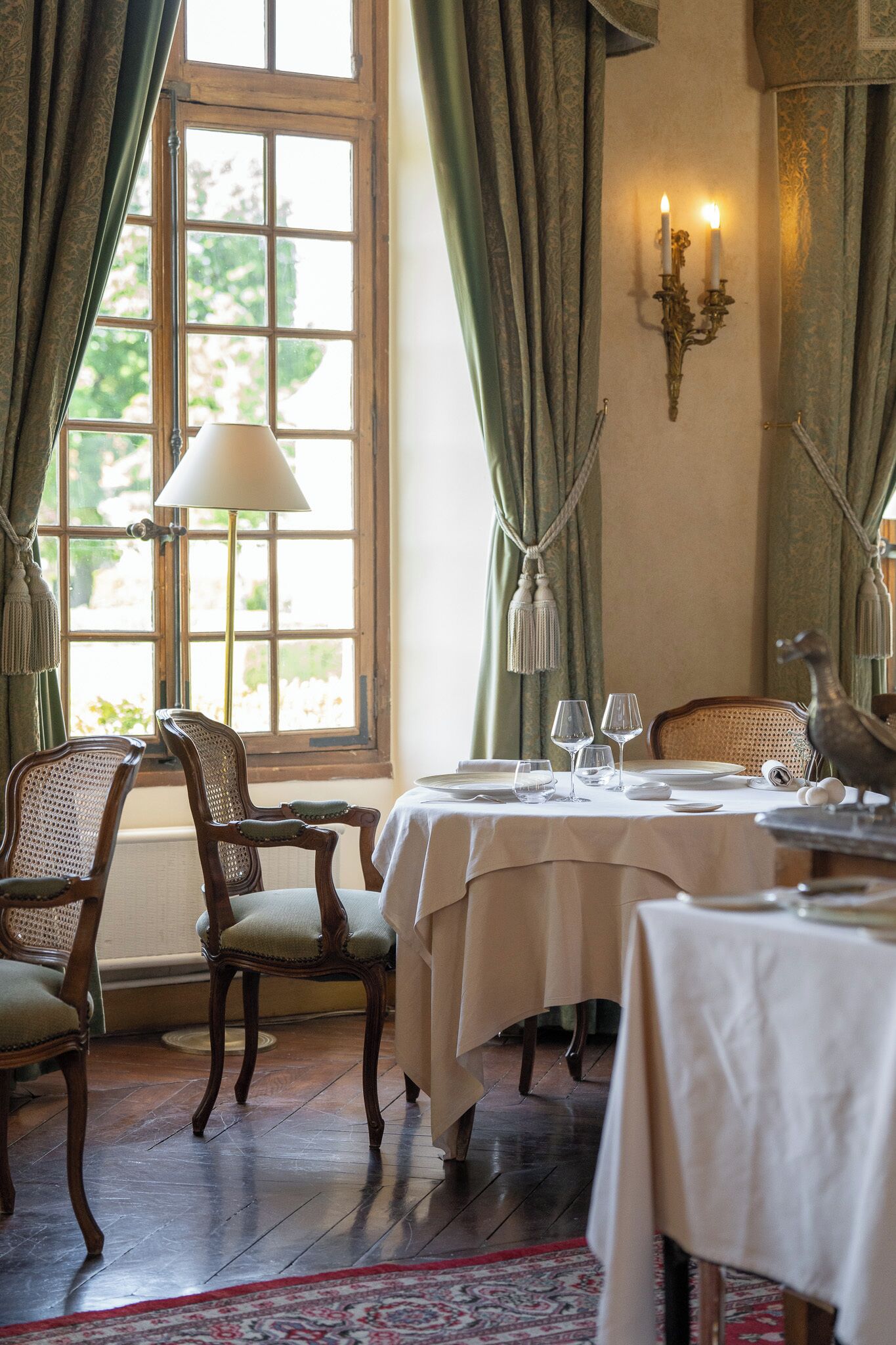 Uma sala de jantar com uma mesa posta para quatro pessoas, duas cadeiras, um abajur e um candelabro fixado na parede.